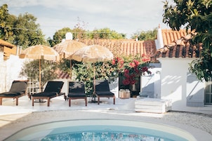 Uma área de piscina com espreguiçadeiras e guarda-sóis de palha, cercada por um muro branco e vegetação.