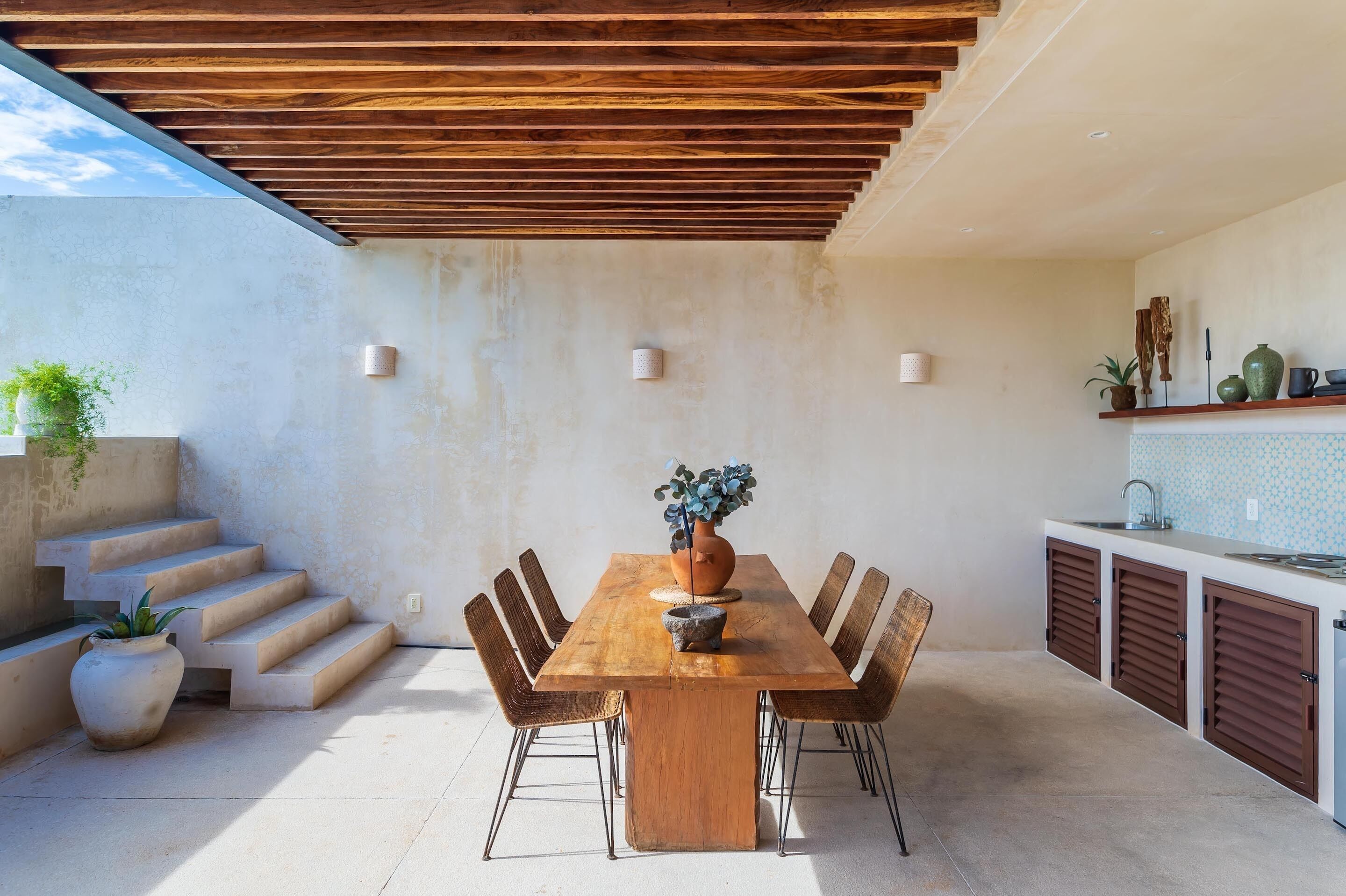 Uma sala de jantar espaçosa com uma mesa e cadeiras de madeira, uma cozinha com revestimento de azulejos e uma escadaria de concreto.