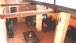Um espaço interno com parede de pedra, teto de madeira e piso de azulejos. Há uma mesa com uma vela, um sofá e uma cadeira.