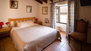 Quarto com cama grande, vigas de madeira no teto, uma cadeira e janela com cortinas.