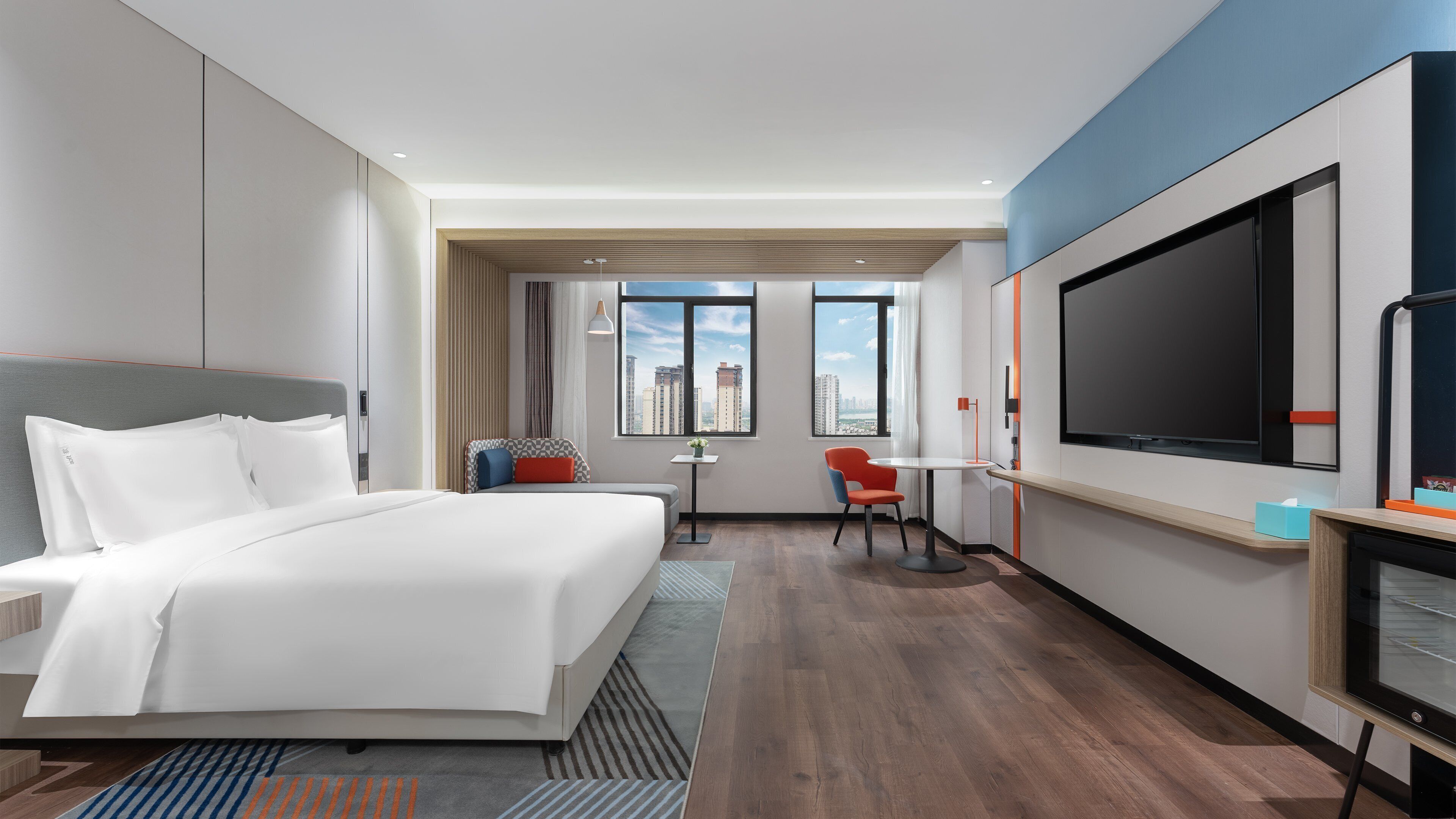 Quarto de hotel com cama, escrivaninha, poltrona, televisão grande e vista para a cidade.