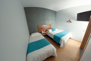 Quarto de hotel moderno com duas camas de solteiro, uma cabeceira de madeira, uma mesa de cabeceira com telefone e uma TV fixada na parede.