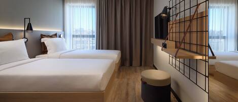 Quarto de hotel moderno com duas camas, uma janela grande com cortinas, piso de madeira e uma prateleira na parede com uma luminária.