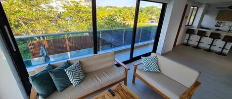Sala de estar moderna com uma janela grande, dois sofás, uma mesa de centro de madeira e uma área de cozinha com banquetas.