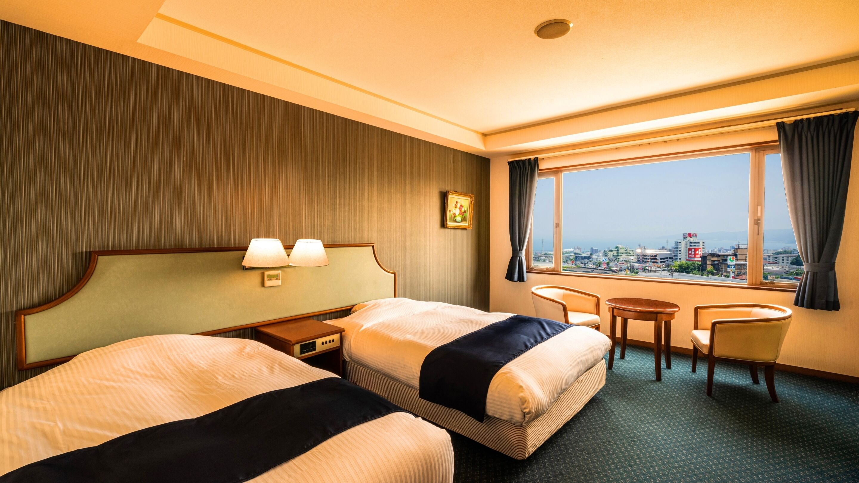 Quarto de hotel com duas camas, uma mesa pequena, duas cadeiras, uma janela com vista para a cidade e uma luminária fixada na parede.