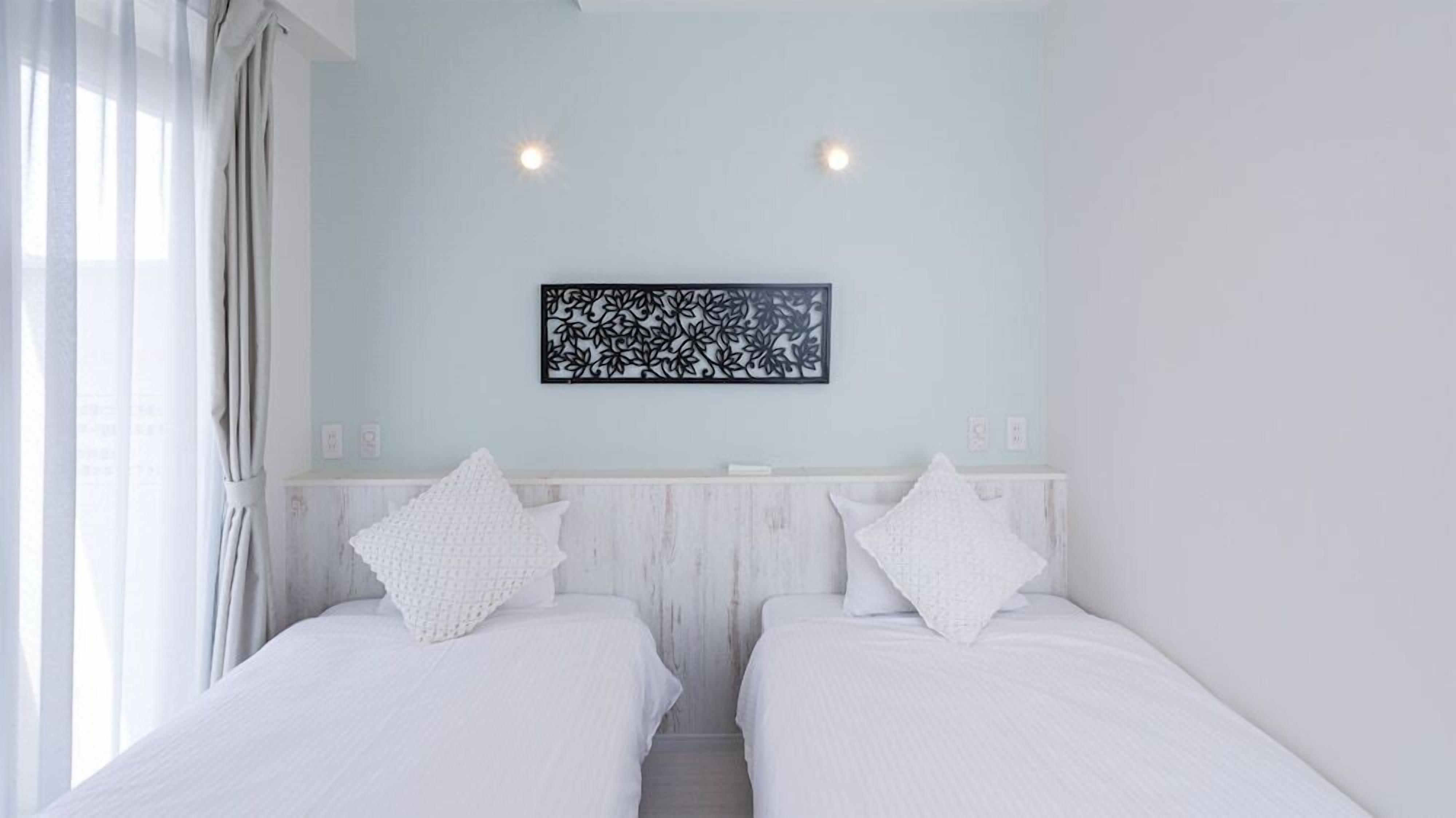 Quarto com duas camas, parede azul clara, painel preto decorativo e janela com cortinas.