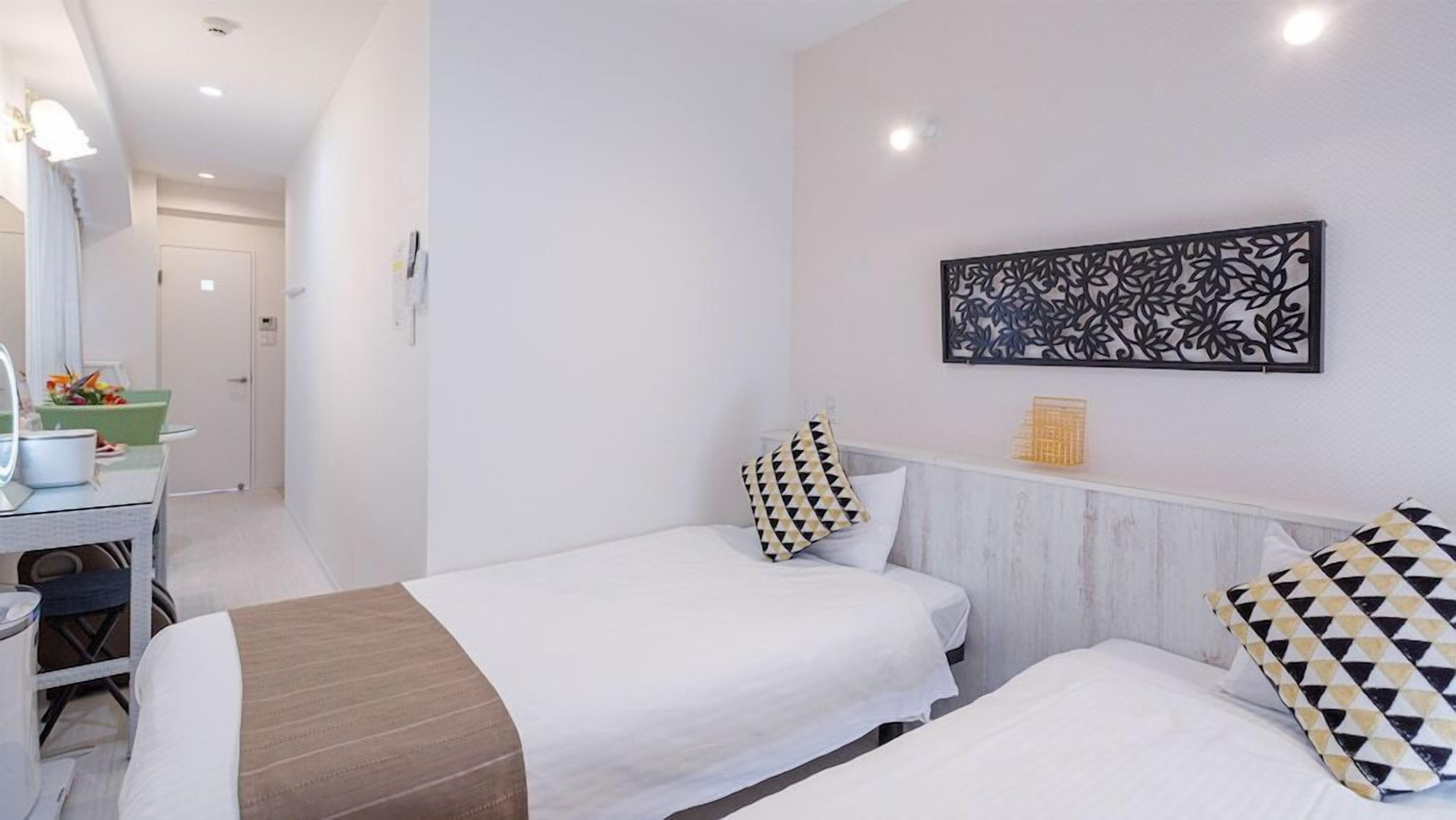 Quarto de hotel com duas camas, uma escrivaninha com espelho, uma mesinha com uma cesta de frutas e um objeto decorativo na parede.