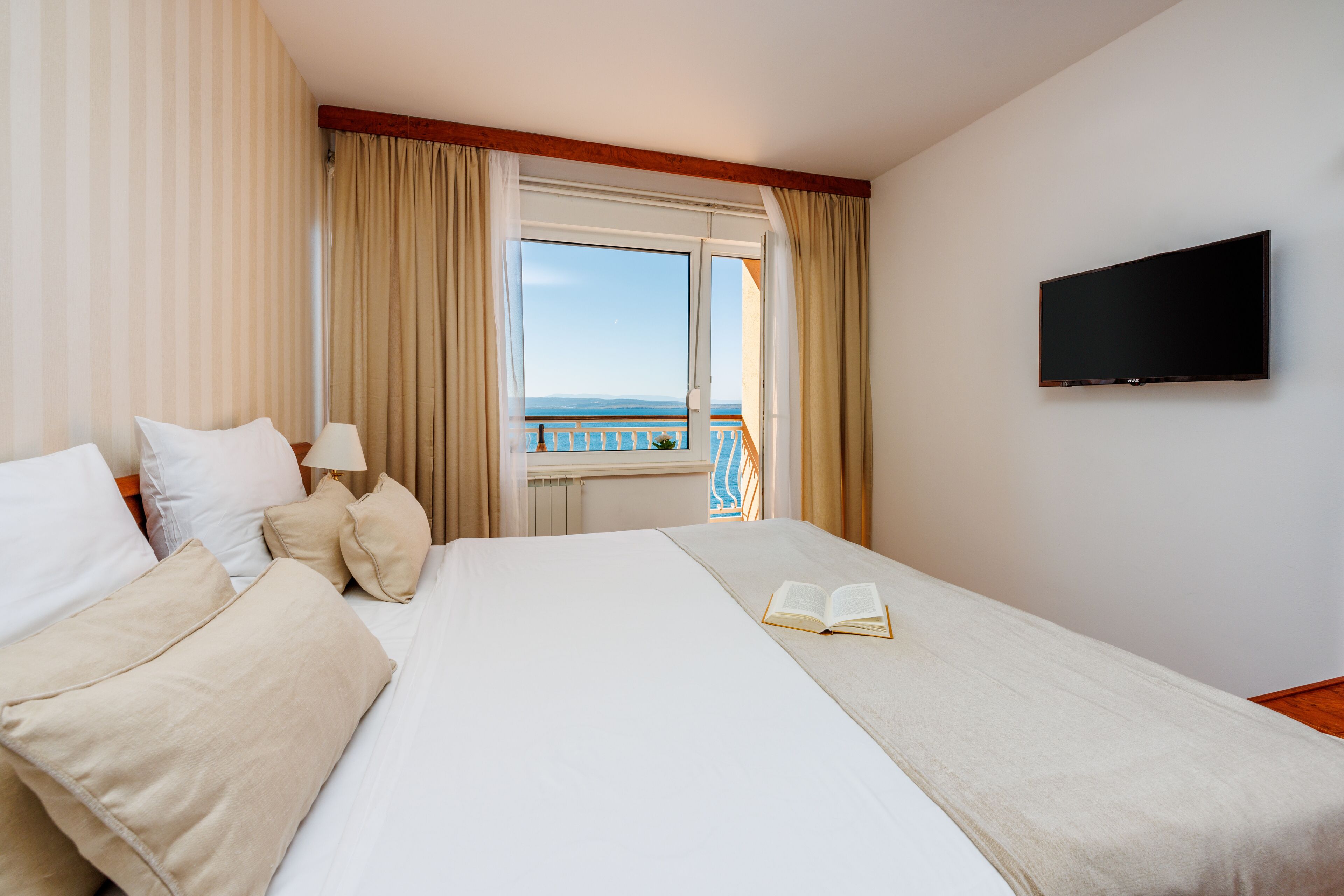 Quarto de hotel com uma cama grande, televisão presa na parede e vista para o mar pela janela.