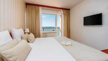 Quarto de hotel com uma cama grande, televisão presa na parede e vista para o mar pela janela.