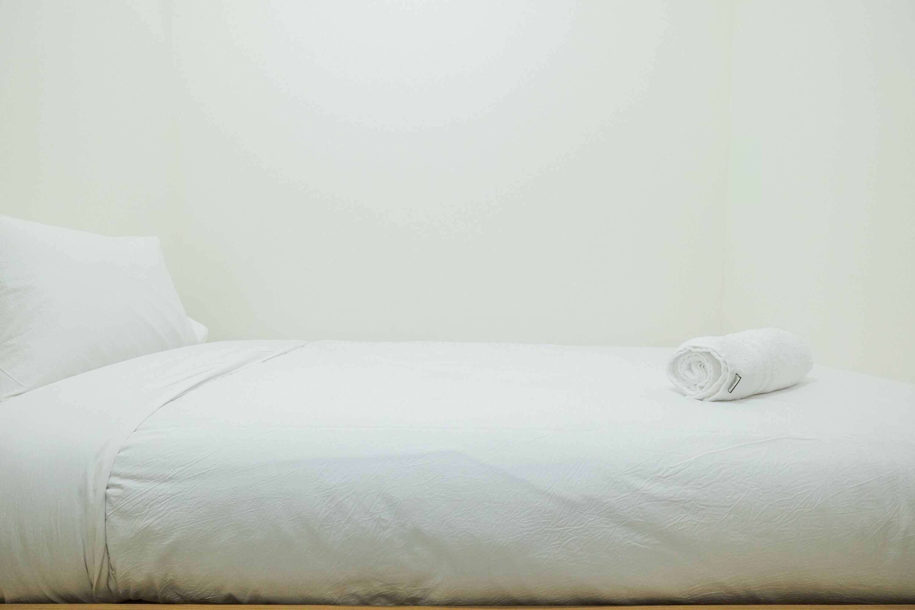 Cama bem arrumada com uma toalha branca enrolada por cima, um travesseiro branco e uma parede lisa ao fundo.