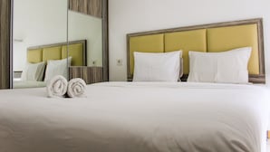 Cama bem arrumada com lençóis brancos e duas toalhas enroladas, cabeceira de madeira e um espelho refletindo parte do quarto.