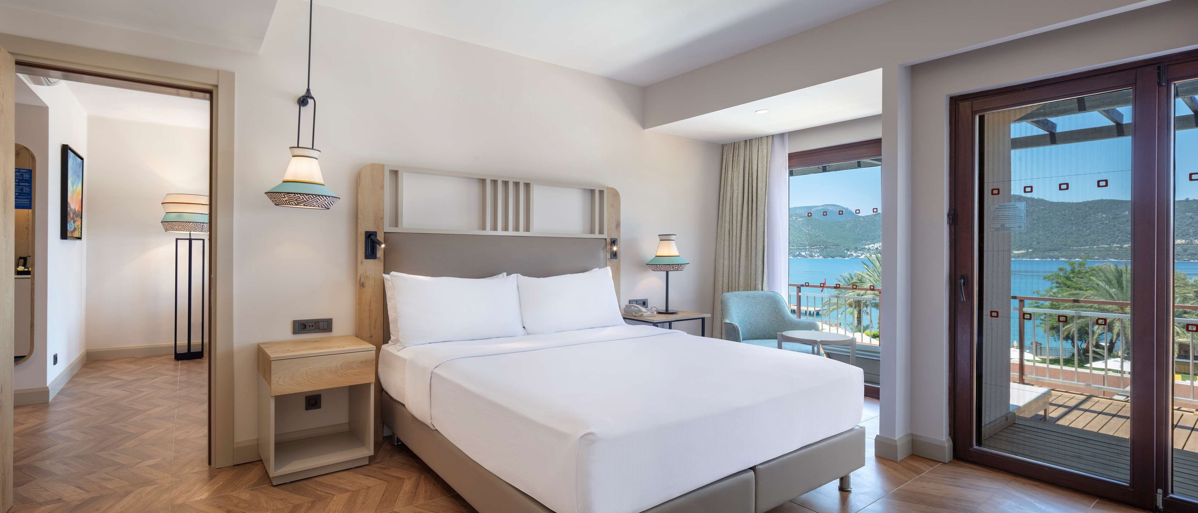 Quarto de hotel com cama, mesa de cabeceira, luminária e vista para o mar.