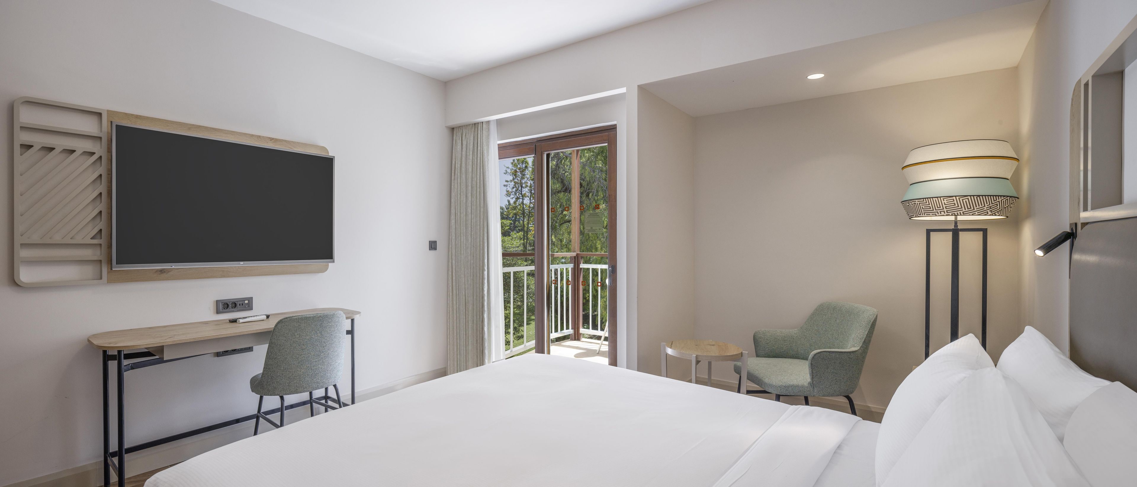 Quarto de hotel com cama, escrivaninha com cadeira, televisão, varanda com cadeiras e abajur.