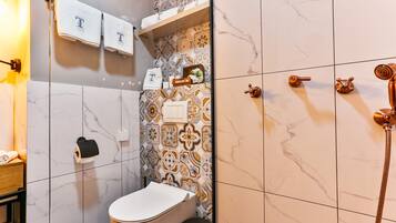 Casa de banho com sanita, um porta-toalhas com toalhas, um suporte para papel higiénico, chuveiro com torneira de cobre e parede com azulejos estampados.