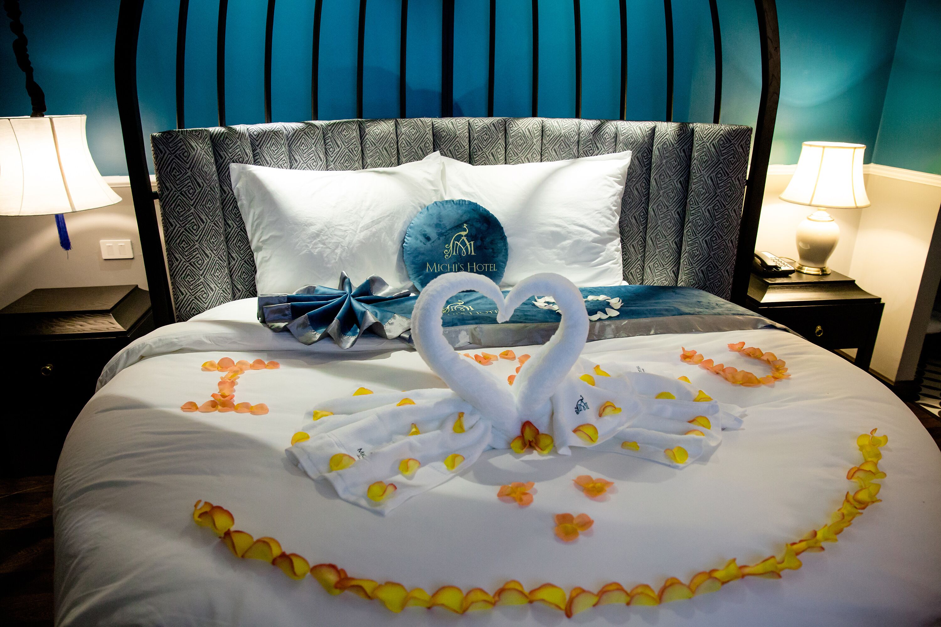 Cama bem arrumada com arranjo de toalha em formato de coração e pétalas de flores, travesseiro com logotipo de hotel e cabeceira com padrão geométrico.