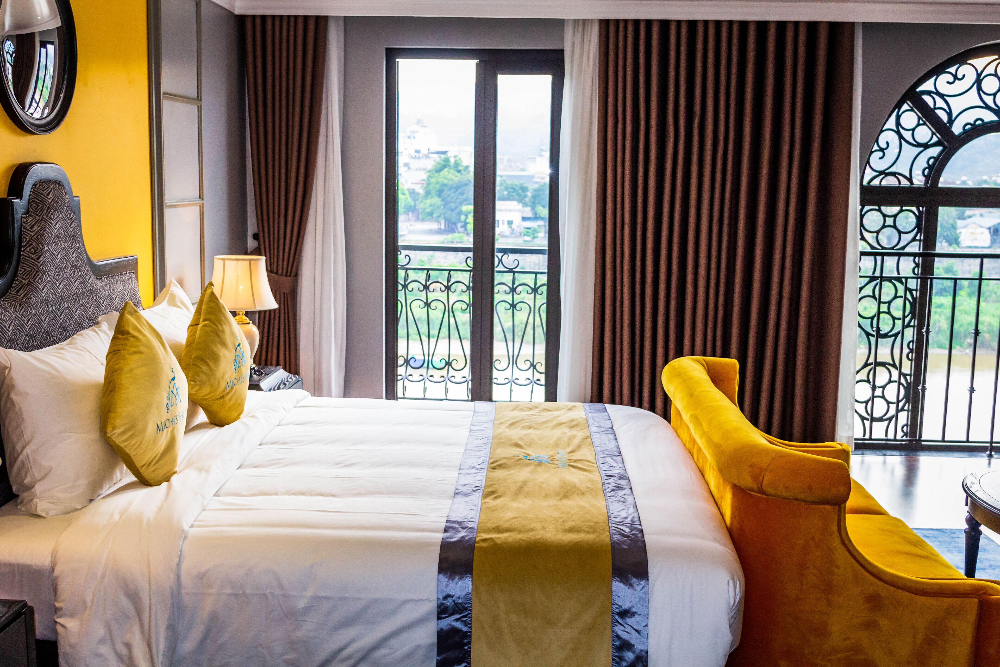 Quarto de hotel com cama, mesa de cabeceira, luminária, cadeira e vista para o exterior pela janela.