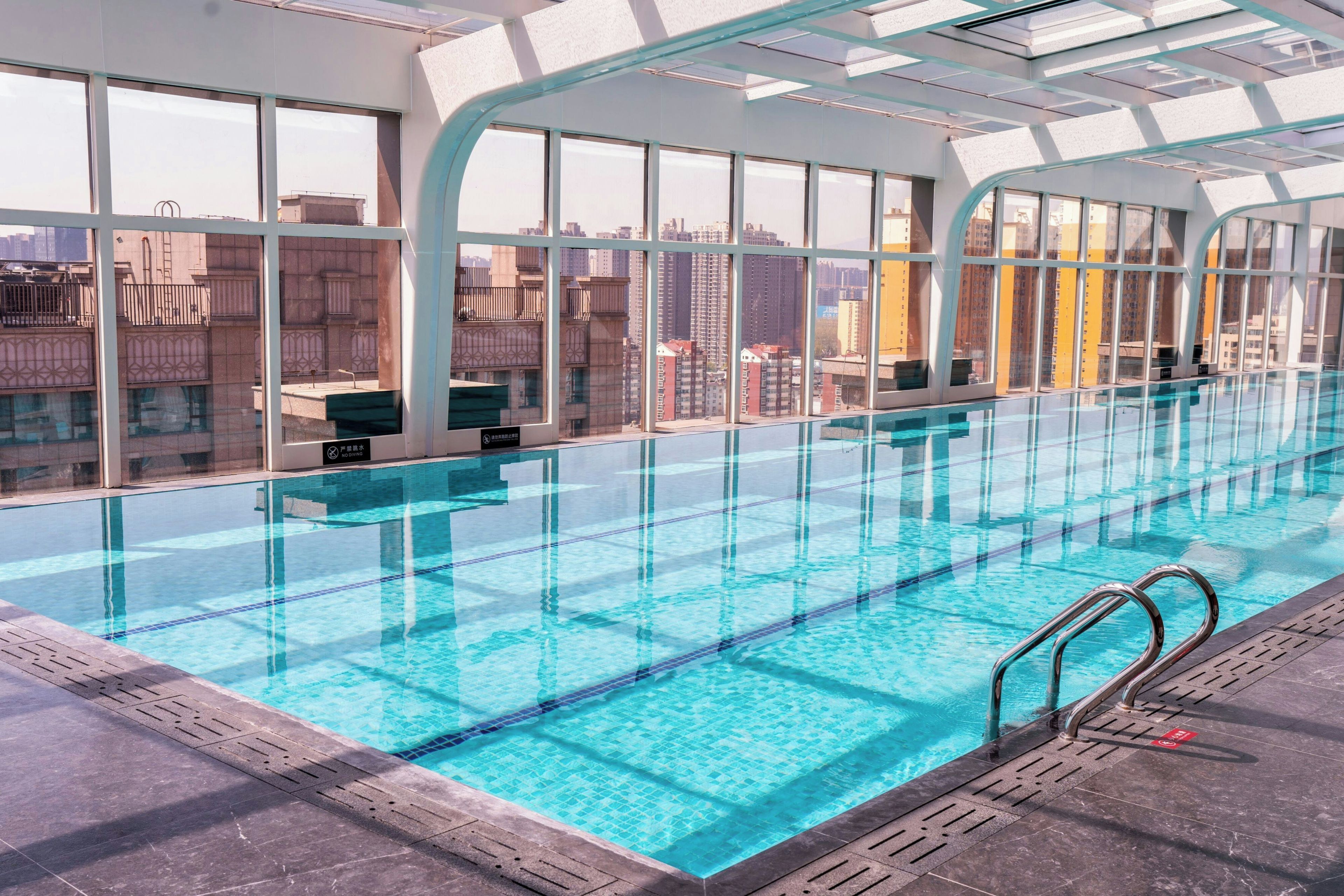 Piscina interior moderna com água azul cristalina, rodeada por amplas janelas com vista para a cidade.