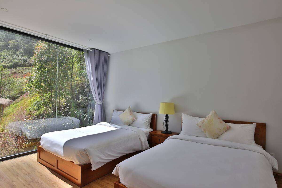 Quarto com duas camas, uma mesinha de cabeceira, uma luminária e uma janela grande com vista para áreas verdes.