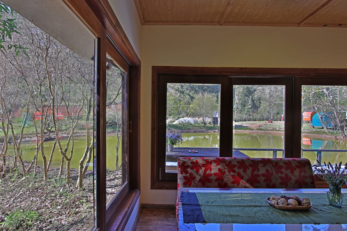 Quarto com uma janela ampla que dá para um lago e árvores. Há um sofá com uma almofada floral e uma mesa com uma tigela de frutas e um vaso.