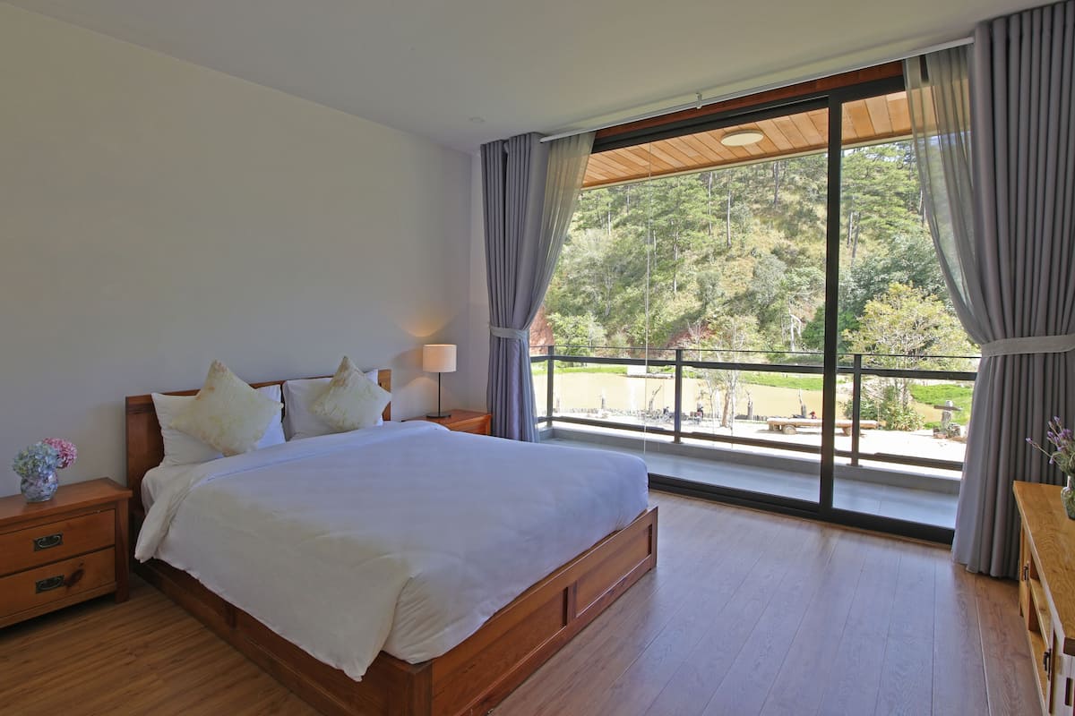 Quarto com cama grande, cabeceira de madeira, mesas de cabeceira e vista para uma floresta através de uma janela ampla.