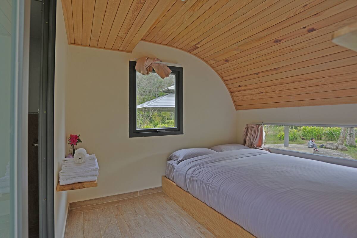 Quarto com teto de madeira, janela com vista para vegetação, cama com colchão branco e uma pequena estante com toalhas dobradas.