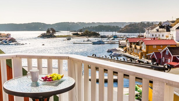 Varanda com mesa, um jarro e uma tigela de frutas, com vista para uma marina com barcos e edifícios.