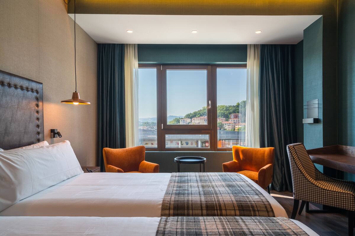 Quarto de hotel com uma cama grande, duas poltronas, uma pequena mesa e vista para a cidade através de amplas janelas.
