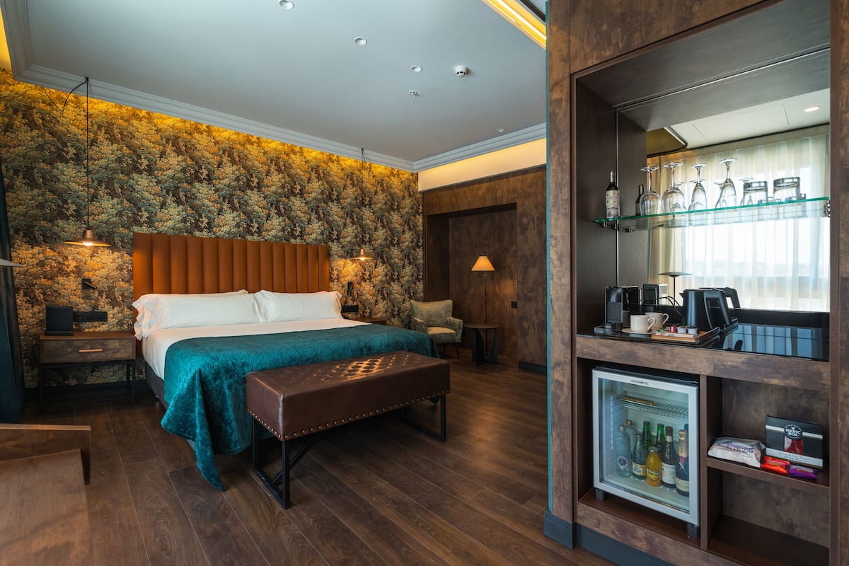Quarto de hotel moderno com uma cama grande, um banco, minibar e um espaço de estar com uma cadeira e mesa.