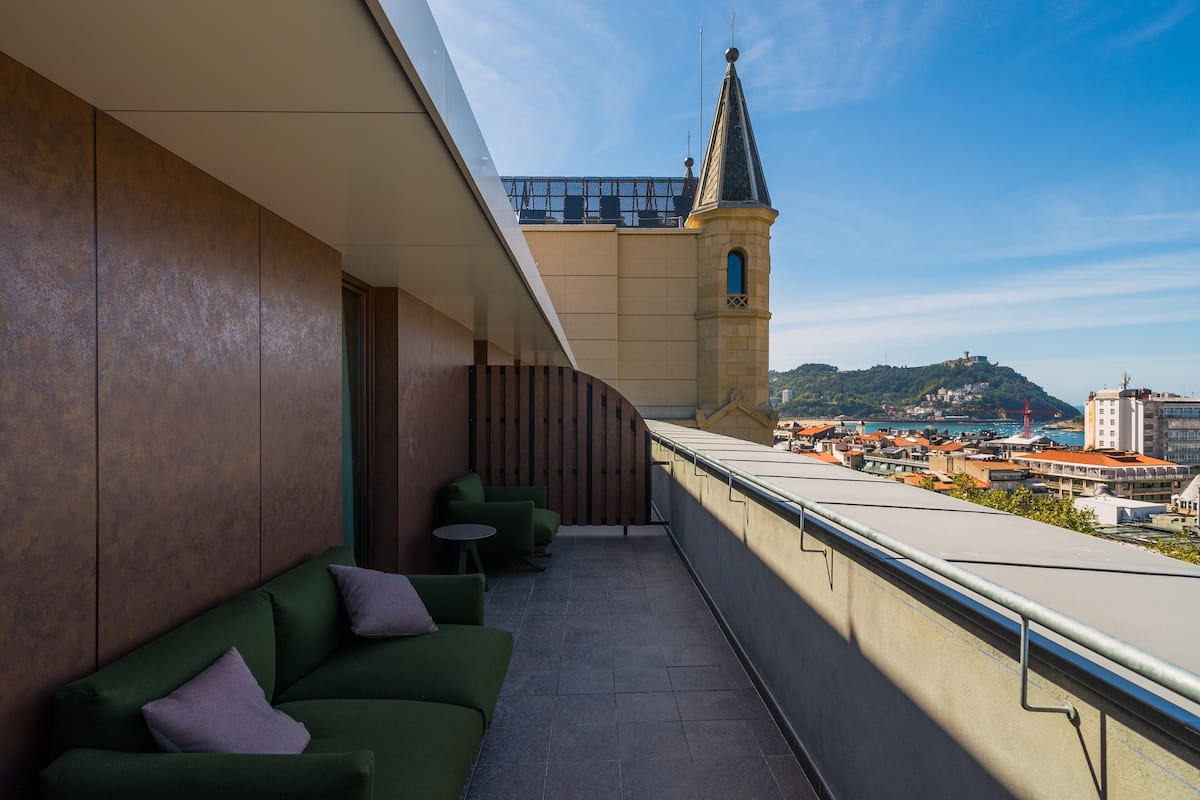 Uma varanda com sofá verde e mesa redonda pequena, com vista para um horizonte urbano que inclui uma torre de igreja e colinas.
