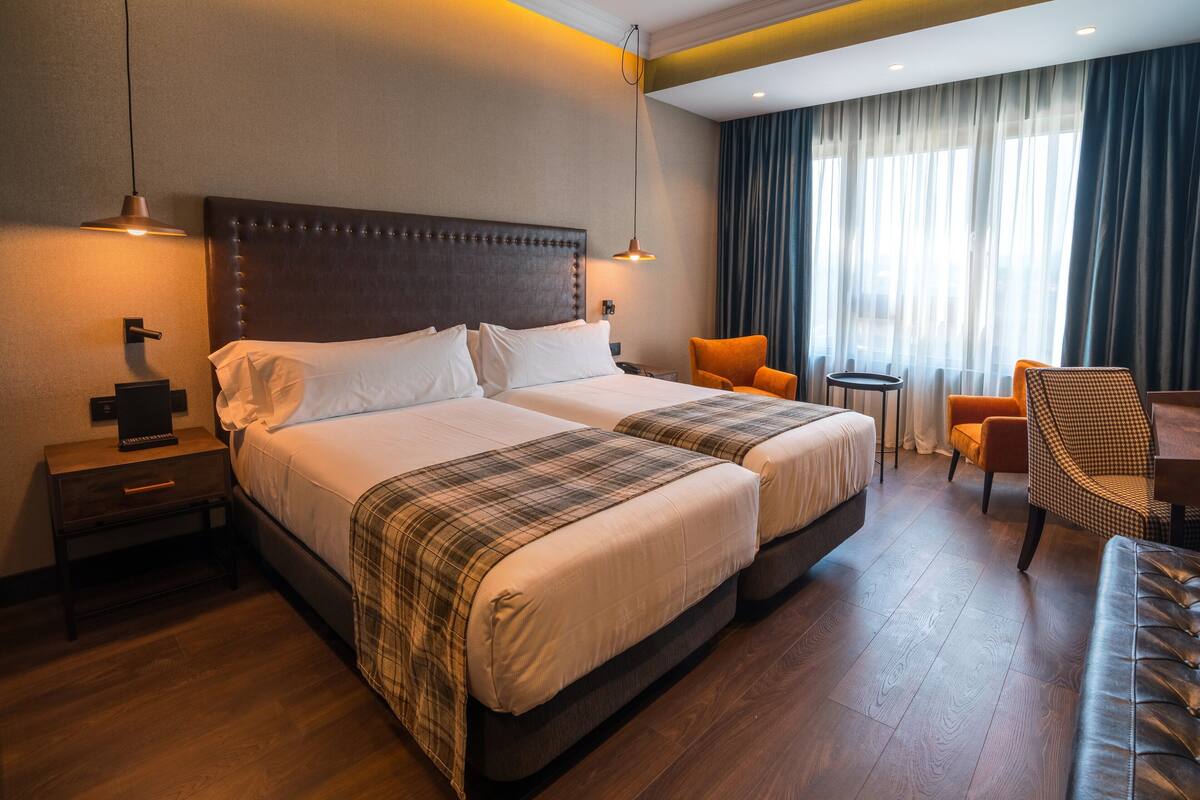 Quarto de hotel com duas camas, uma escrivaninha, uma cadeira e uma mesinha.