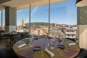 Uma área de refeições com uma mesa redonda grande posta para seis, grandes janelas com vista para a cidade e um edifício alto e ornamentado ao fundo.