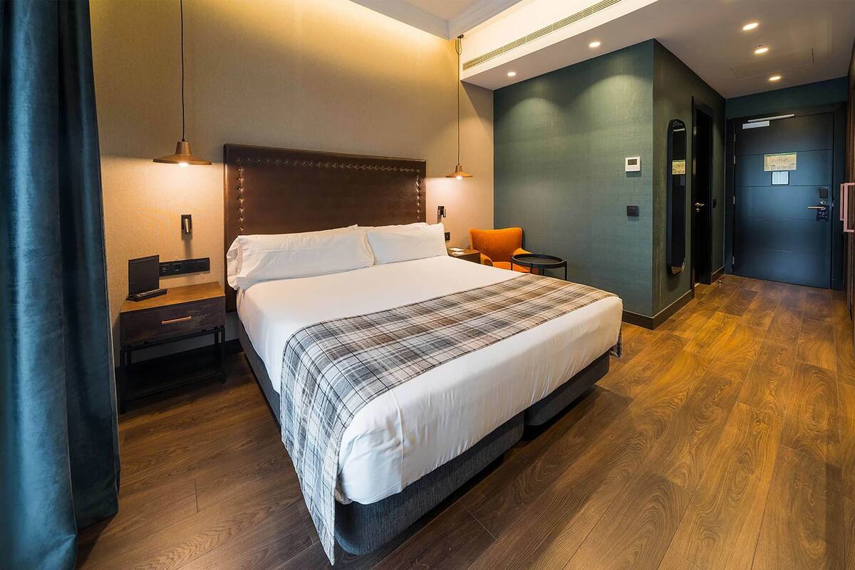 Quarto de hotel com uma cama grande, uma mesa de cabeceira, uma cadeira e uma porta.
