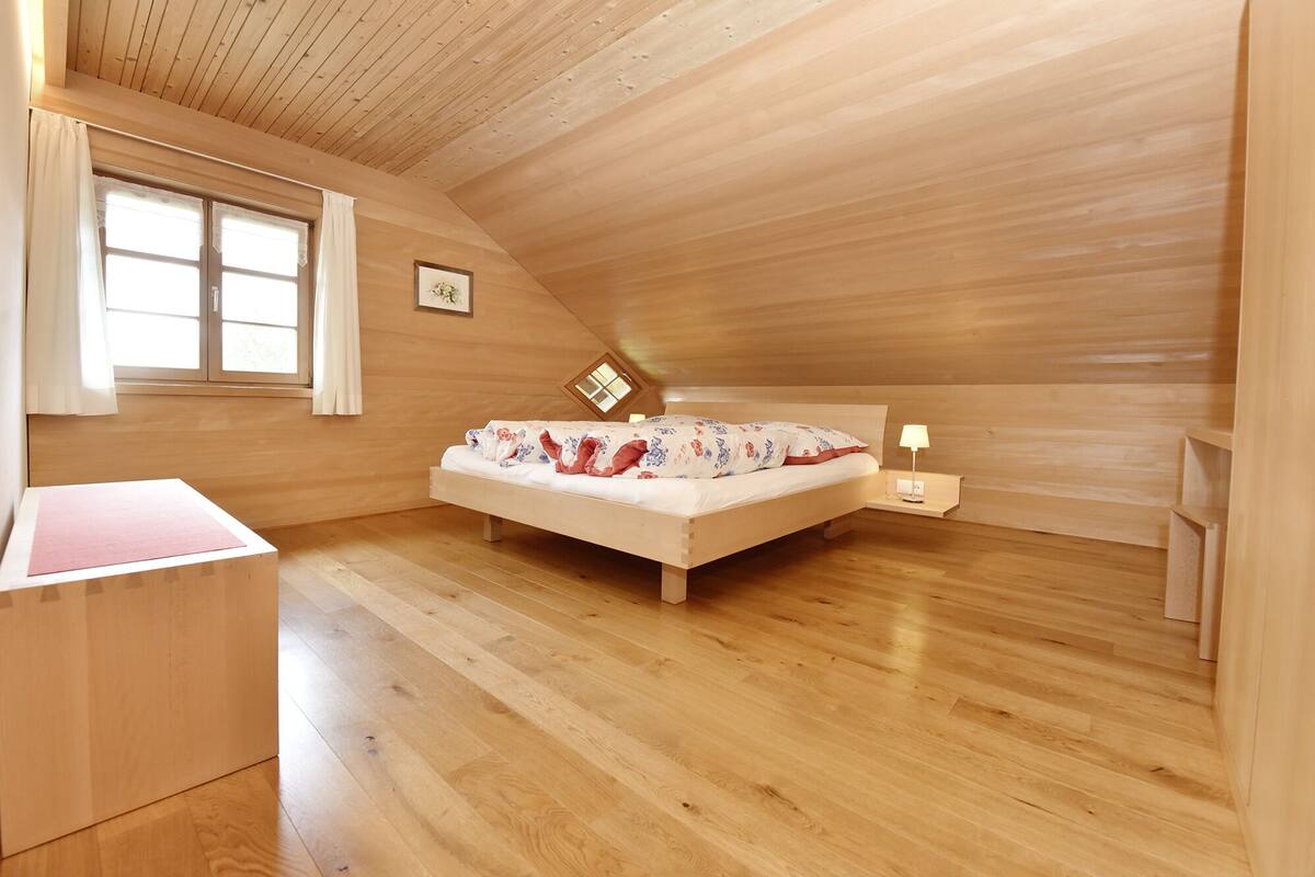 Um quarto com teto de madeira inclinado, uma cama com roupa de cama floral, uma mesa de cabeceira com abajur e uma janela com cortinas.