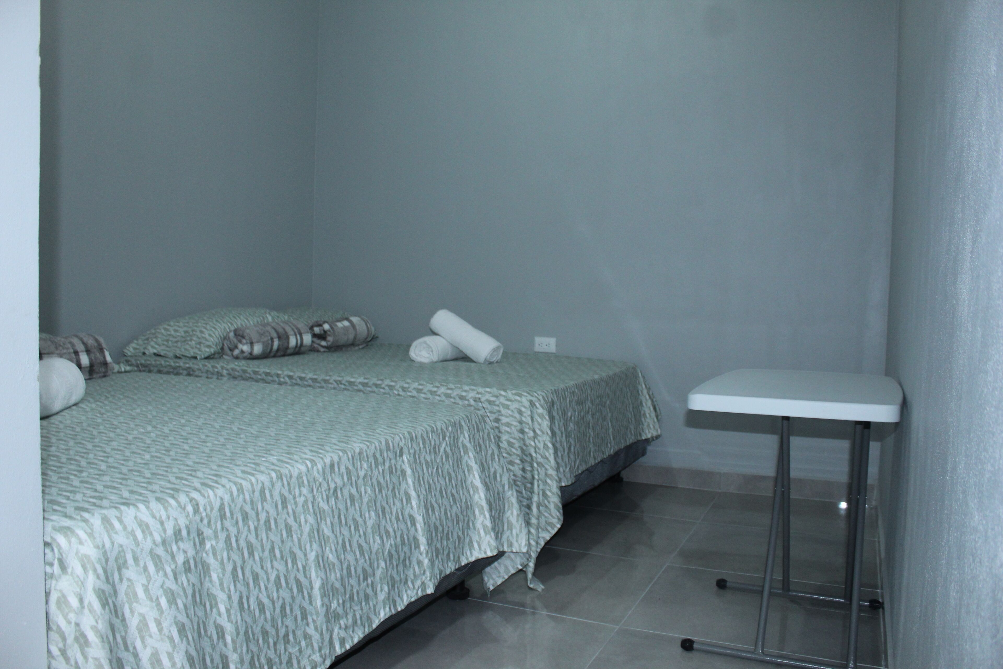 Quarto com cama, almofadas, um cobertor enrolado e um banquinho branco.