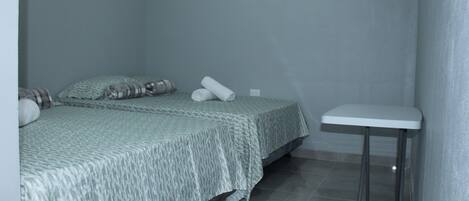 Quarto com cama, almofadas, um cobertor enrolado e um banquinho branco.