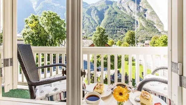 Uma varanda com uma cadeira de vime, uma mesa com comida e bebidas, e vista para as montanhas.