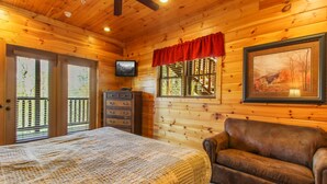 Quarto de cabana de madeira com uma cama, uma televisão, uma cômoda, uma janela com valança vermelha e um quadro na parede.