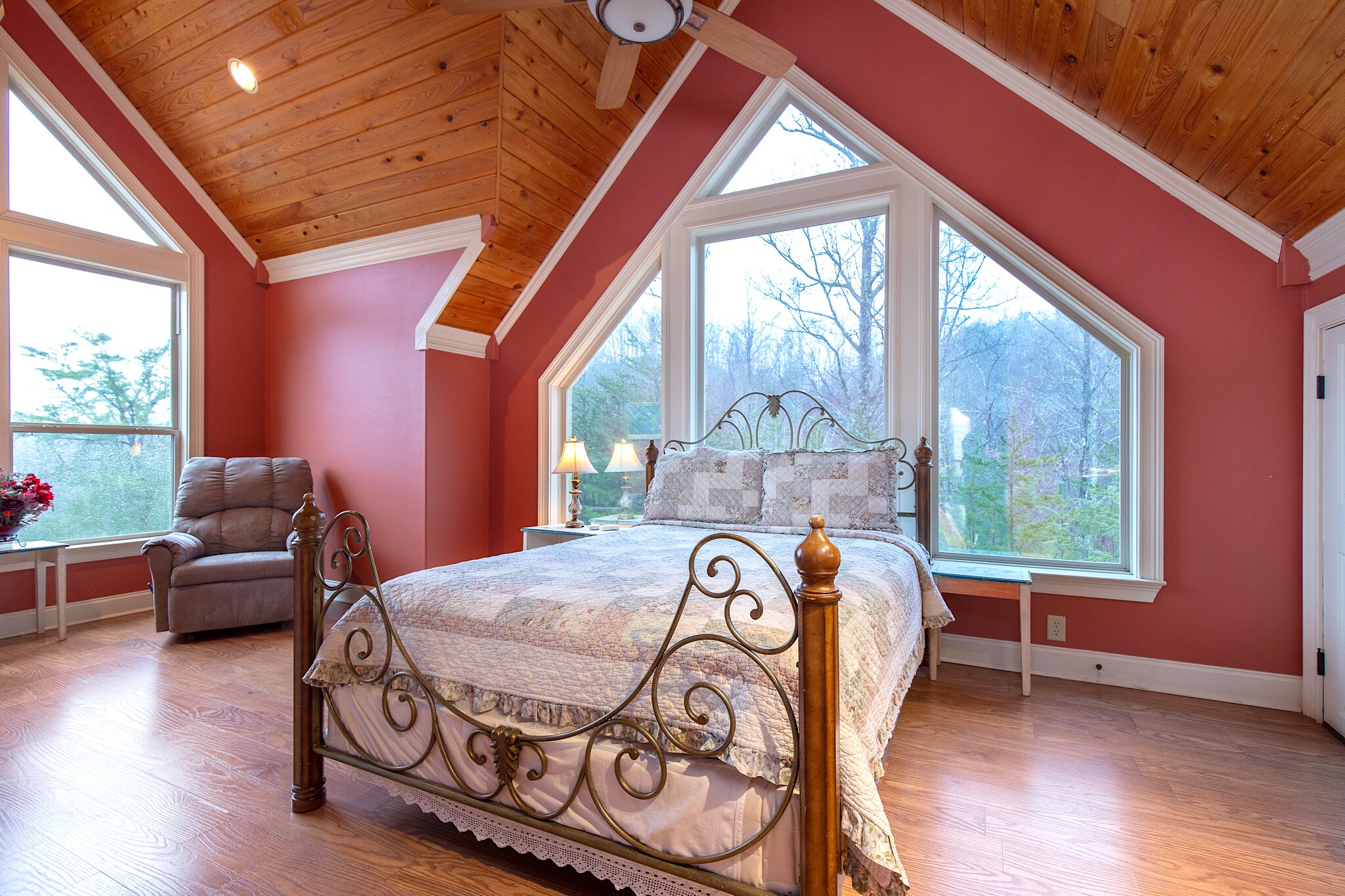 Quarto com teto de madeira, grandes janelas, cama com estrutura metálica, uma cadeira e mesa de cabeceira com luminárias.