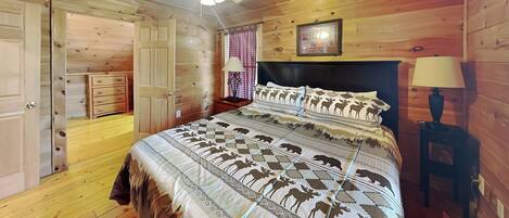 Quarto de cabana de madeira com uma cama, uma mesa de cabeceira, um abajur, um ventilador de teto e uma porta que dá para outro cômodo.