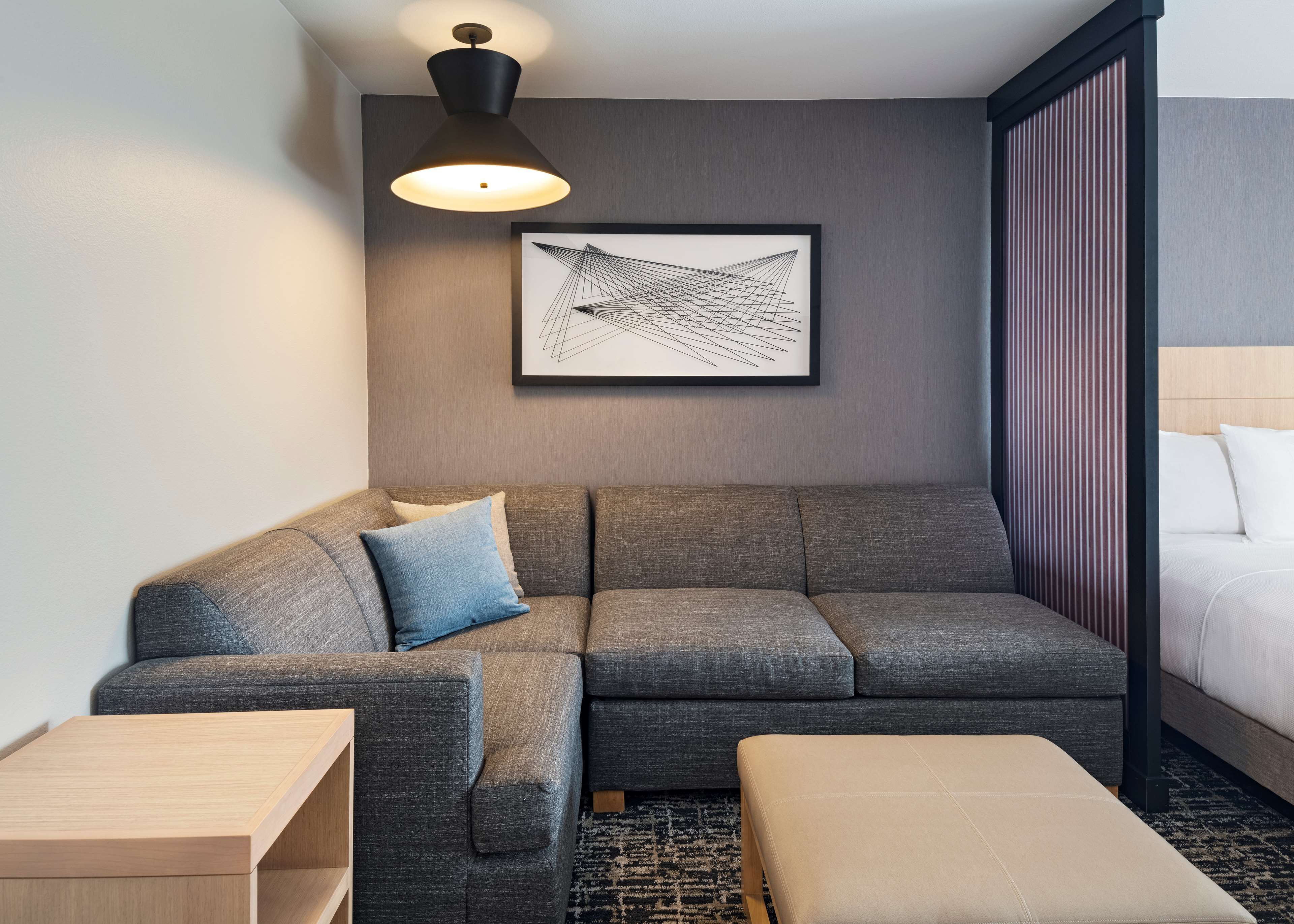 Quarto de hotel moderno com um sofá modular cinza, uma pequena mesa lateral de madeira, um banquinho bege, uma obra de arte abstrata emoldurada na parede e uma cama com roupa de cama branca.