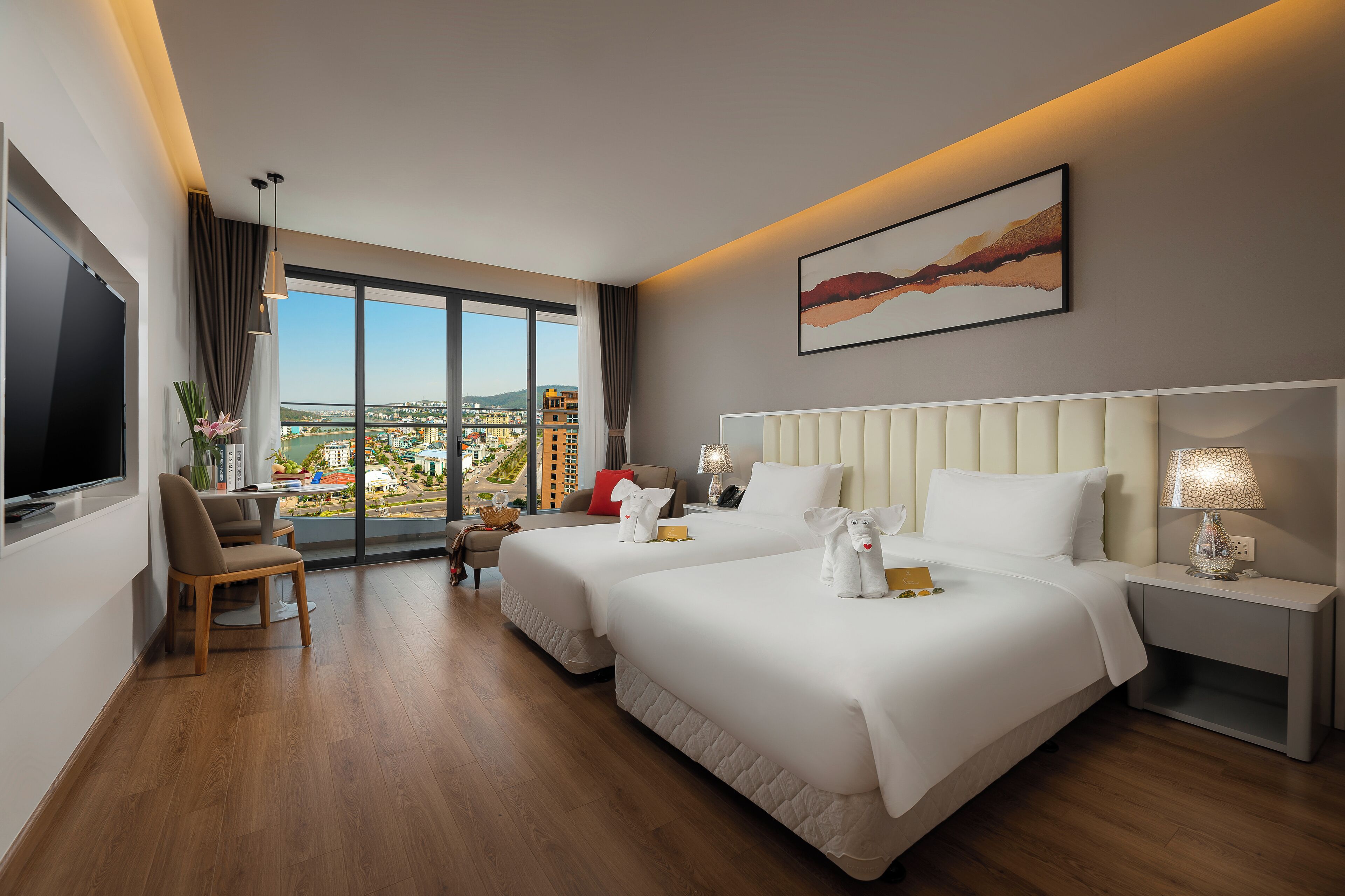 Quarto de hotel com duas camas, televisão, escrivaninha e uma ampla janela com vista para a cidade.