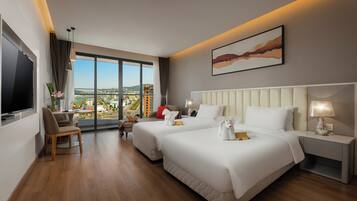 Quarto de hotel com duas camas, televisão, escrivaninha e uma ampla janela com vista para a cidade.