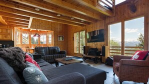 Interior acolhedor de uma cabana de madeira, com sofá, poltronas, televisão e vista panorâmica para as montanhas através de grandes janelas.