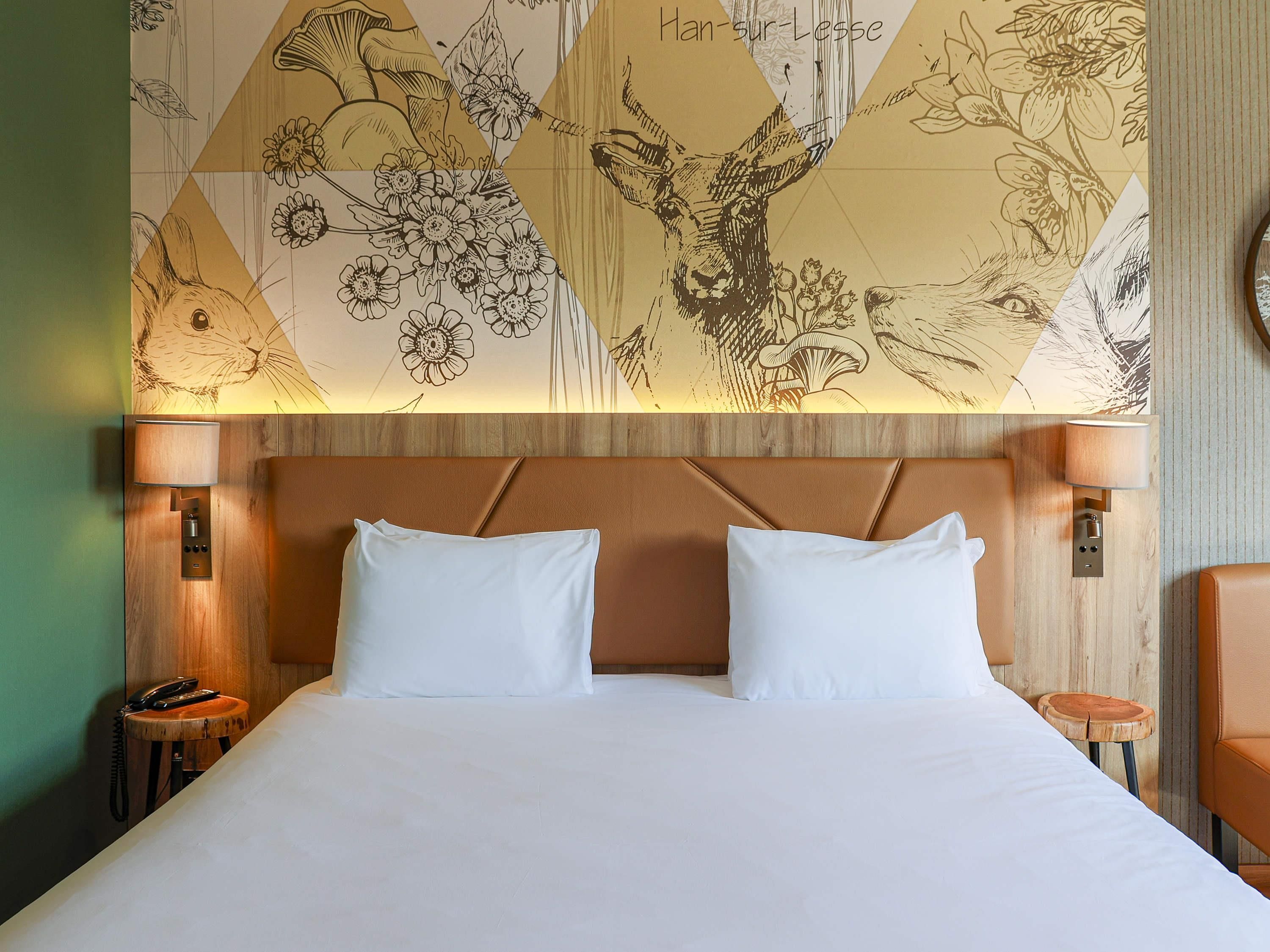 Quarto de hotel com cama, duas almofadas, cabeceira de madeira, parede com padrões florais e abstratos, e duas luminárias de cabeceira.