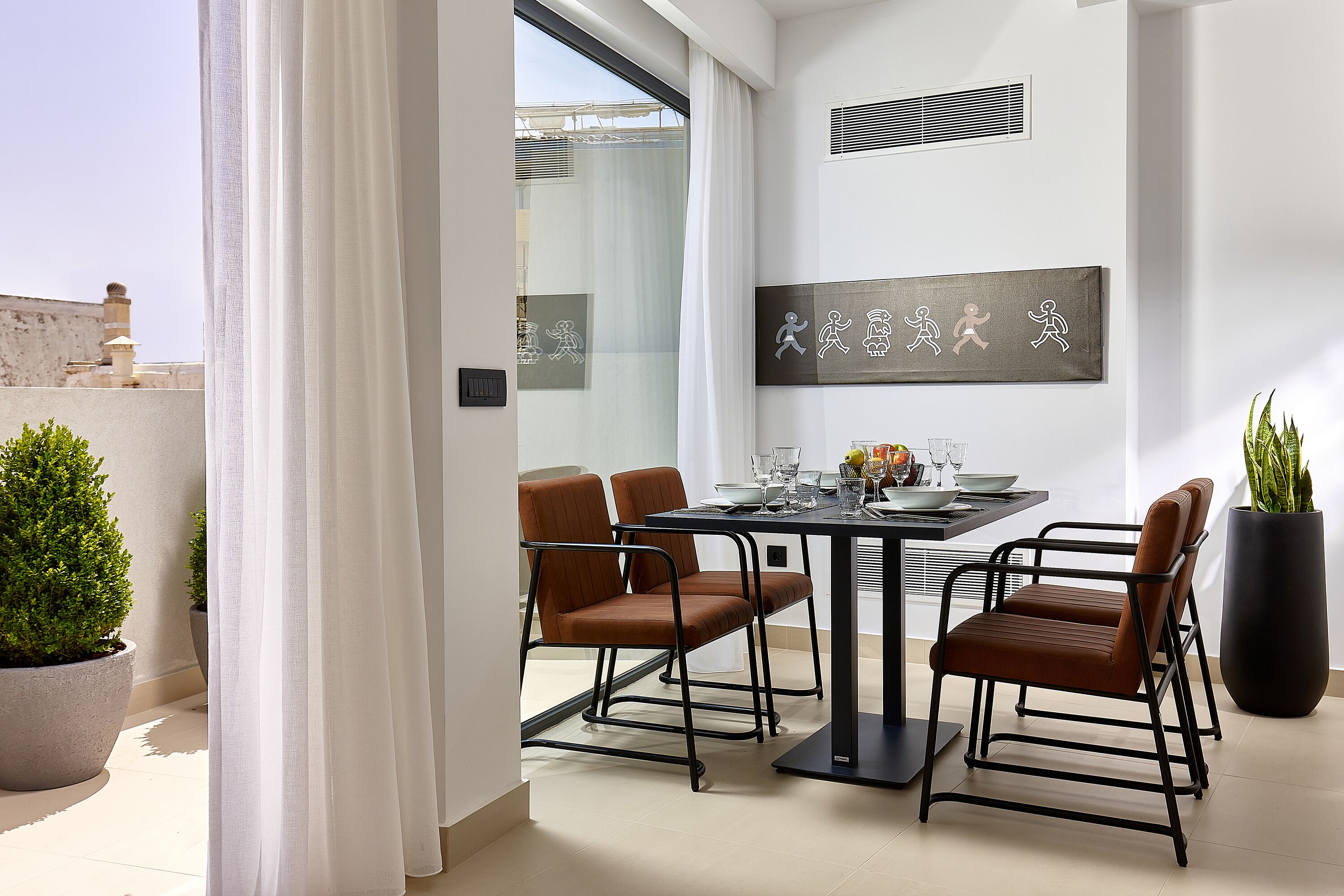 Uma sala de jantar moderna com uma mesa posta para quatro pessoas, rodeada por cadeiras, um vaso grande com planta e uma obra de arte pendurada na parede.