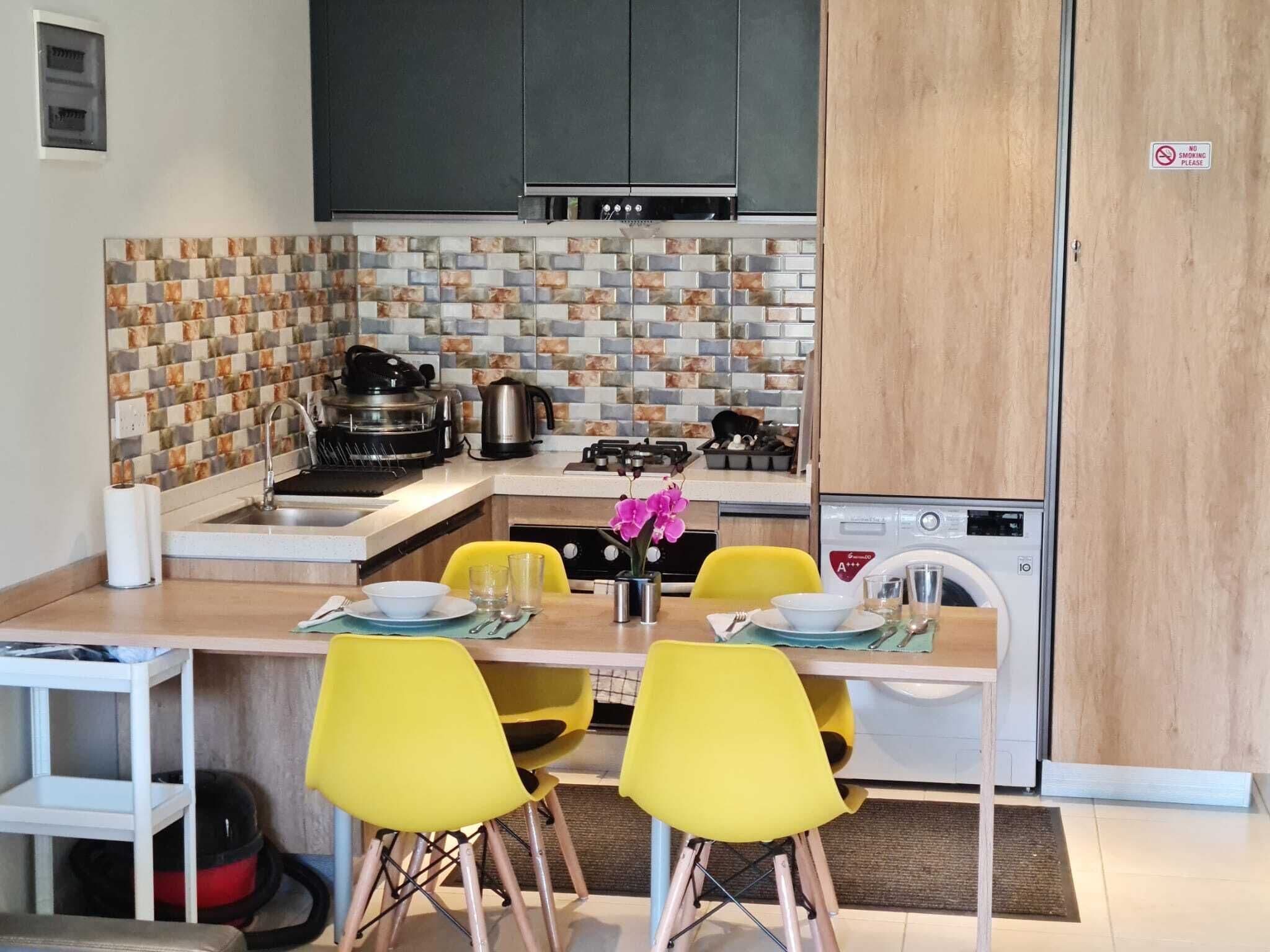 Cozinha compacta com painel de azulejos, fogão a gás, cafeteira e máquina de lavar roupa. Há cadeiras amarelas em torno de uma pequena mesa de jantar.