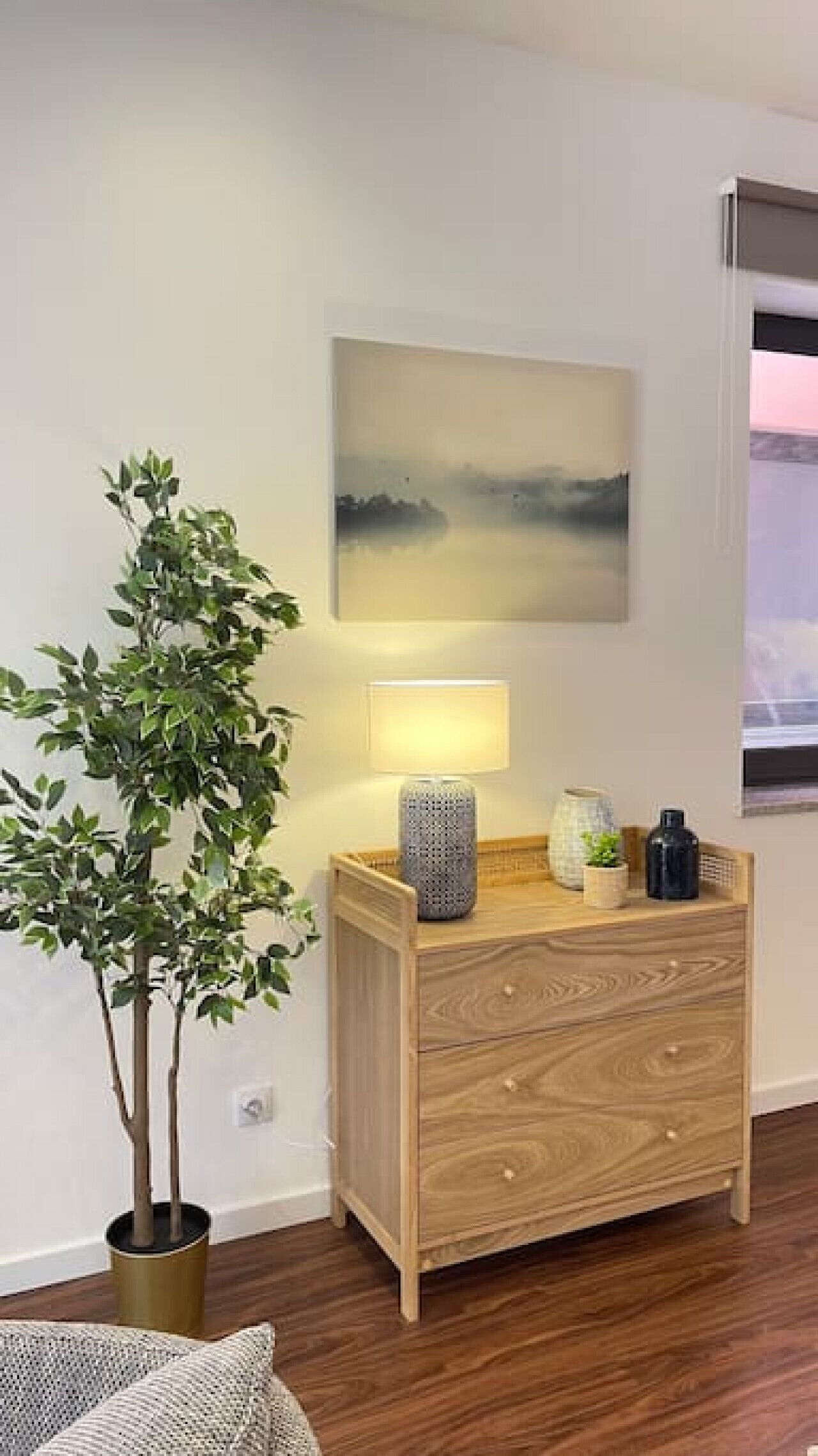 Uma mesa de cabeceira de madeira com uma luminária, um vaso de planta e um quadro na parede.