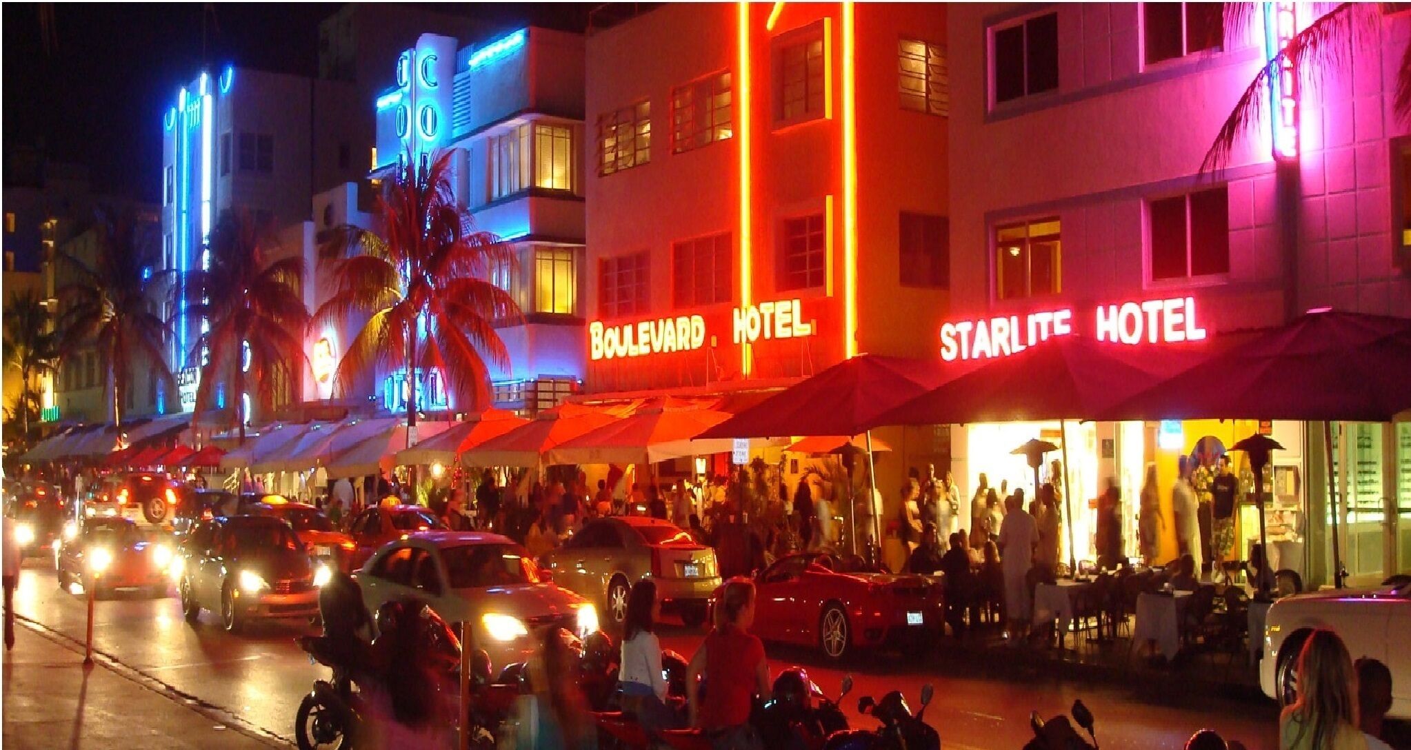 Rua movimentada com dois hotéis, Boulevard Hotel e Starlite Hotel, apresentando letreiros de neon e mesas ao ar livre.