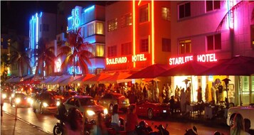 Rua movimentada com dois hotéis, Boulevard Hotel e Starlite Hotel, apresentando letreiros de neon e mesas ao ar livre.