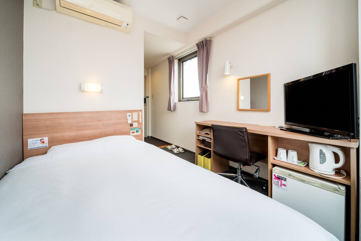Quarto de hotel com cama, escrivaninha com cadeira, televisão, mini-geladeira e janela com cortinas.
